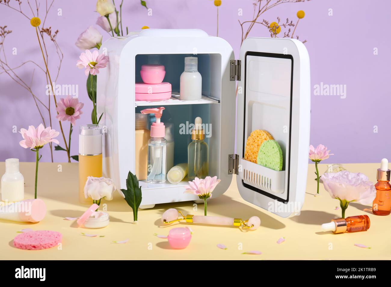 Small refrigerator with cosmetics and flowers on table near lilac wall ...
