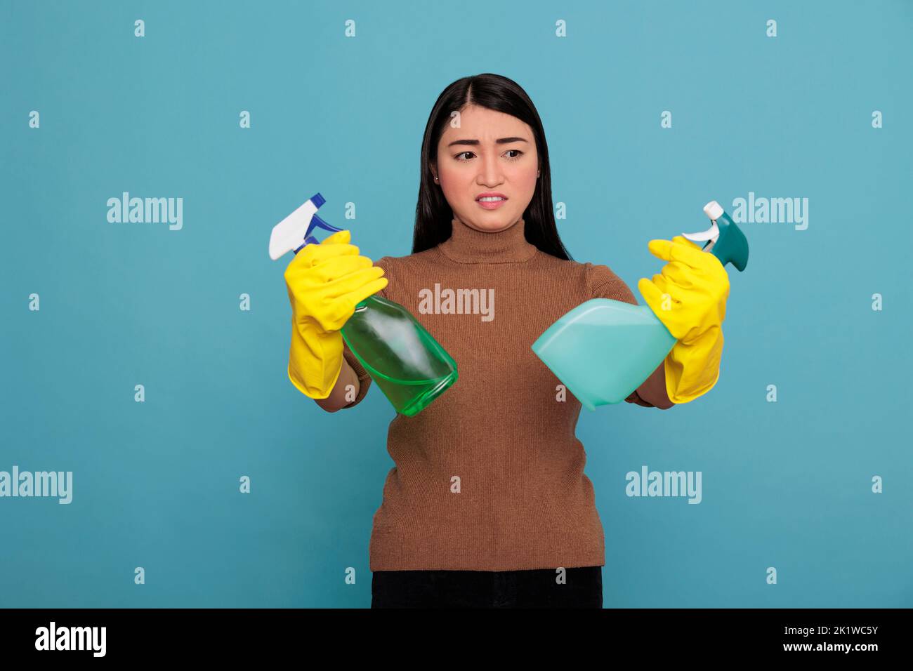 Asian young inactive dull housekeeper from chores holding two detergent ...