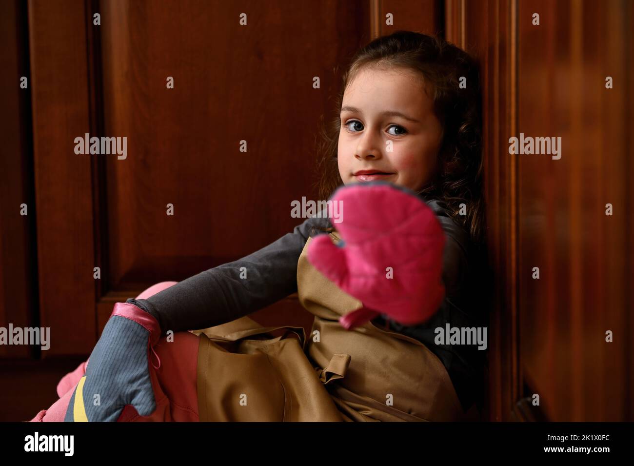 Little girl wearing beige chef's apron, leaning against wooden kitchen ...