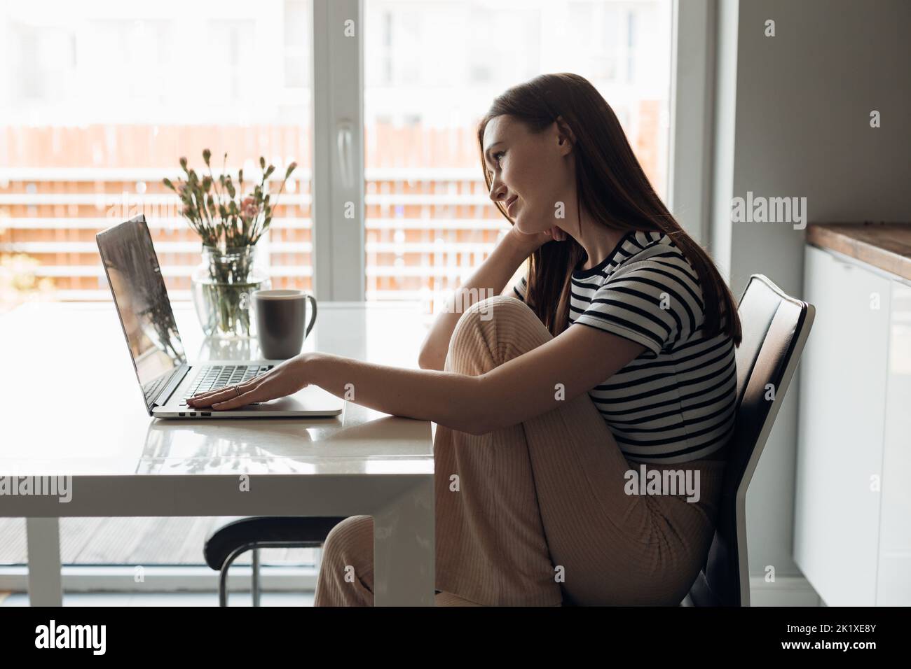 Bored, calm young brown haired woman sitting in the kitchen with bend leg, work on laptop and ...