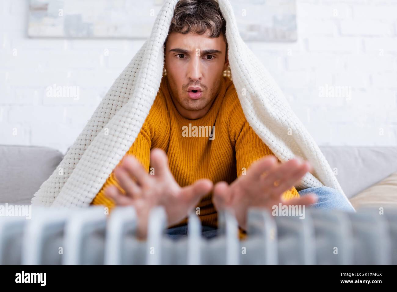 emotional young man covered in blanket warming up hands near radiator ...