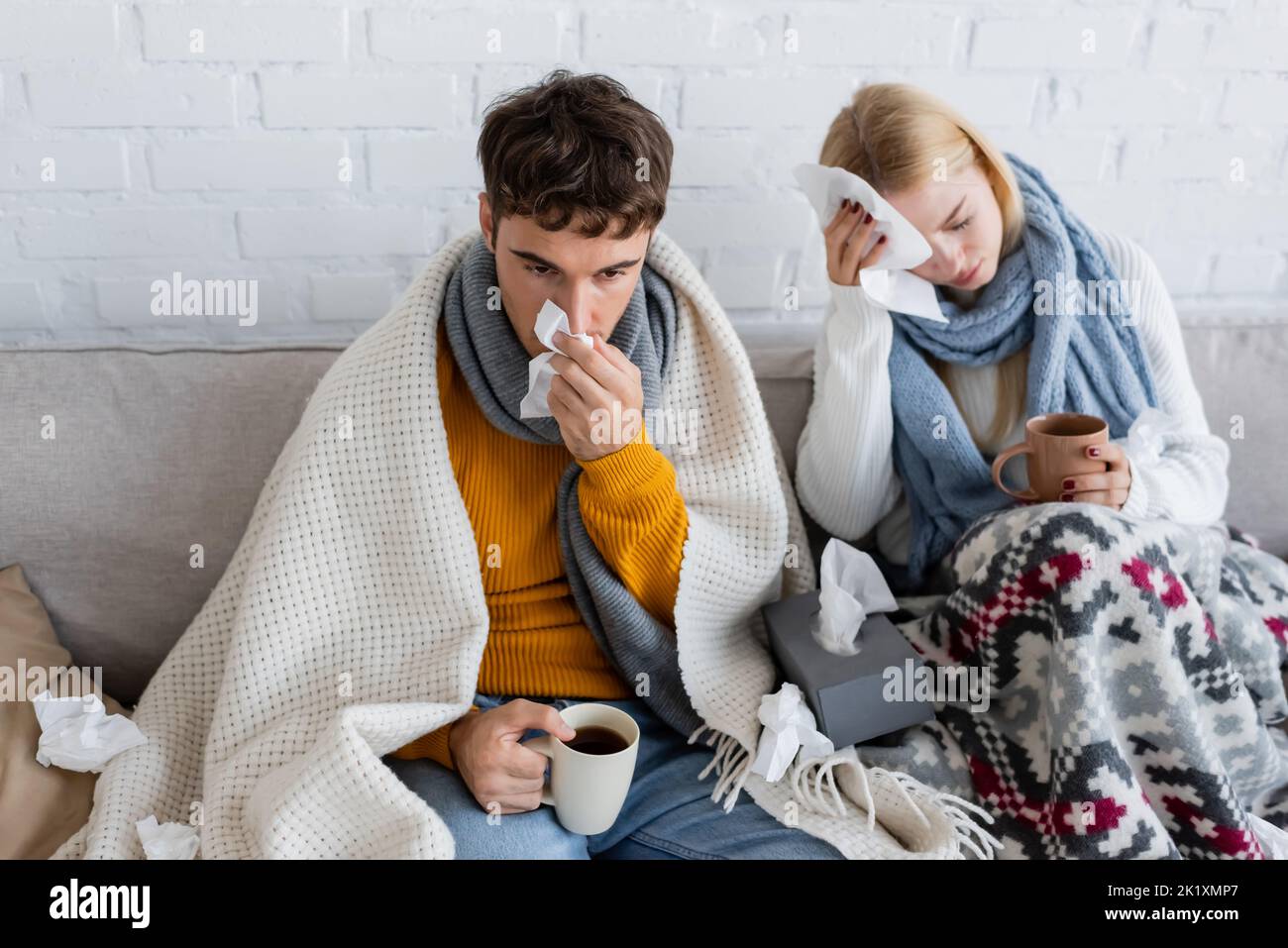 sick couple covered in blankets holding cups of tea and tissues while ...