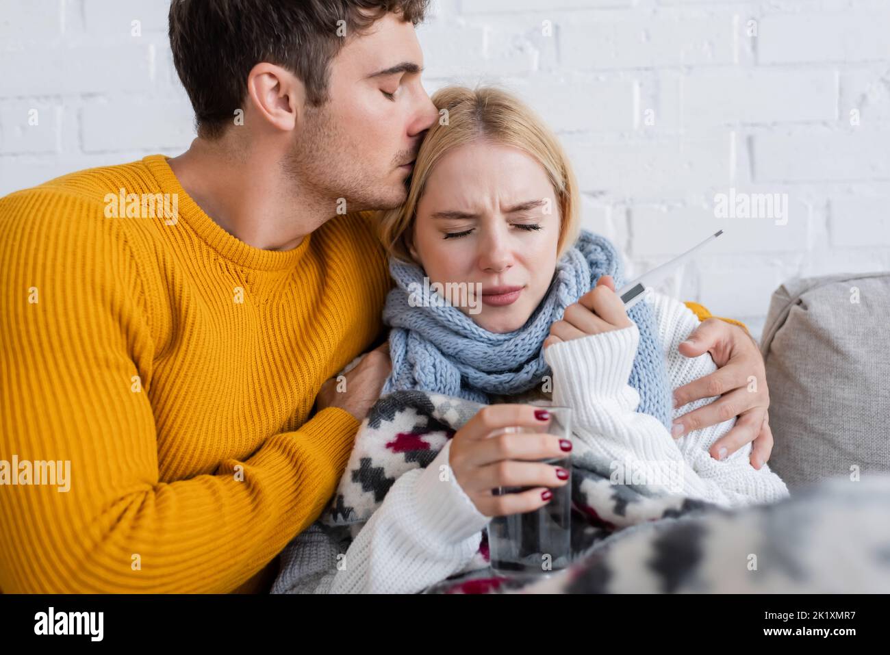 man hugging and kissing head of sick blonde woman with glass of water ...