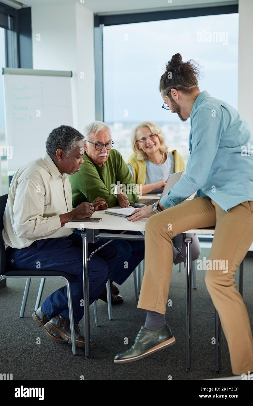 A young hipster teacher is sitting on a desk and explaining to small ...