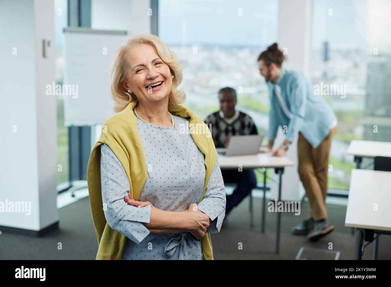 A happy female senior student is standing in a classroom with arms ...