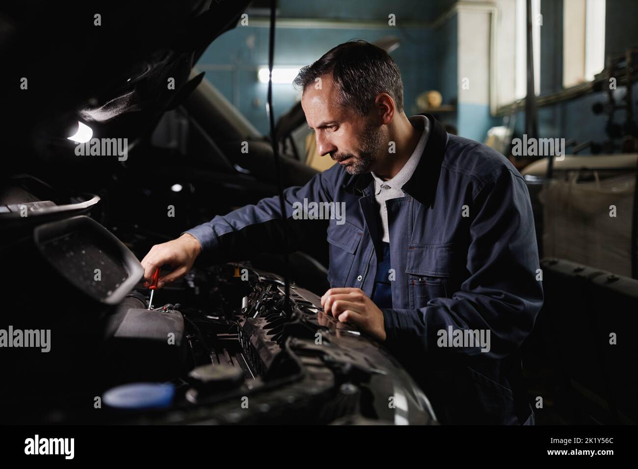 Side view portrait of mature mechanic fixing car engine in dark garage ...