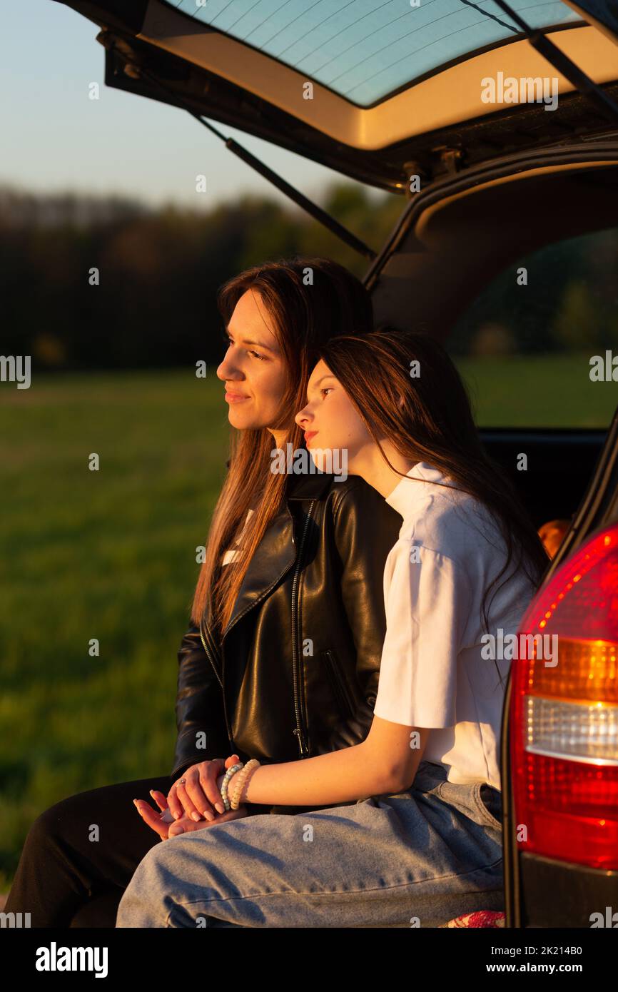 Mother And Daughter Camping On A Hill And Admiring The Sunset While Sitting In The Car Trunk