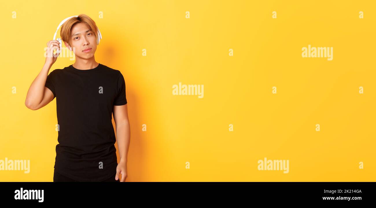 Young handsome asian guy with blond hair, take-off headphones to listen ...