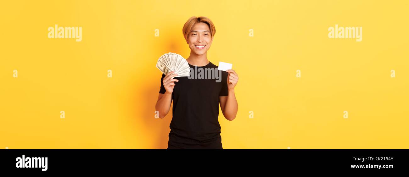 Pleased handsome asian guy showing money and credit card, smiling happy ...