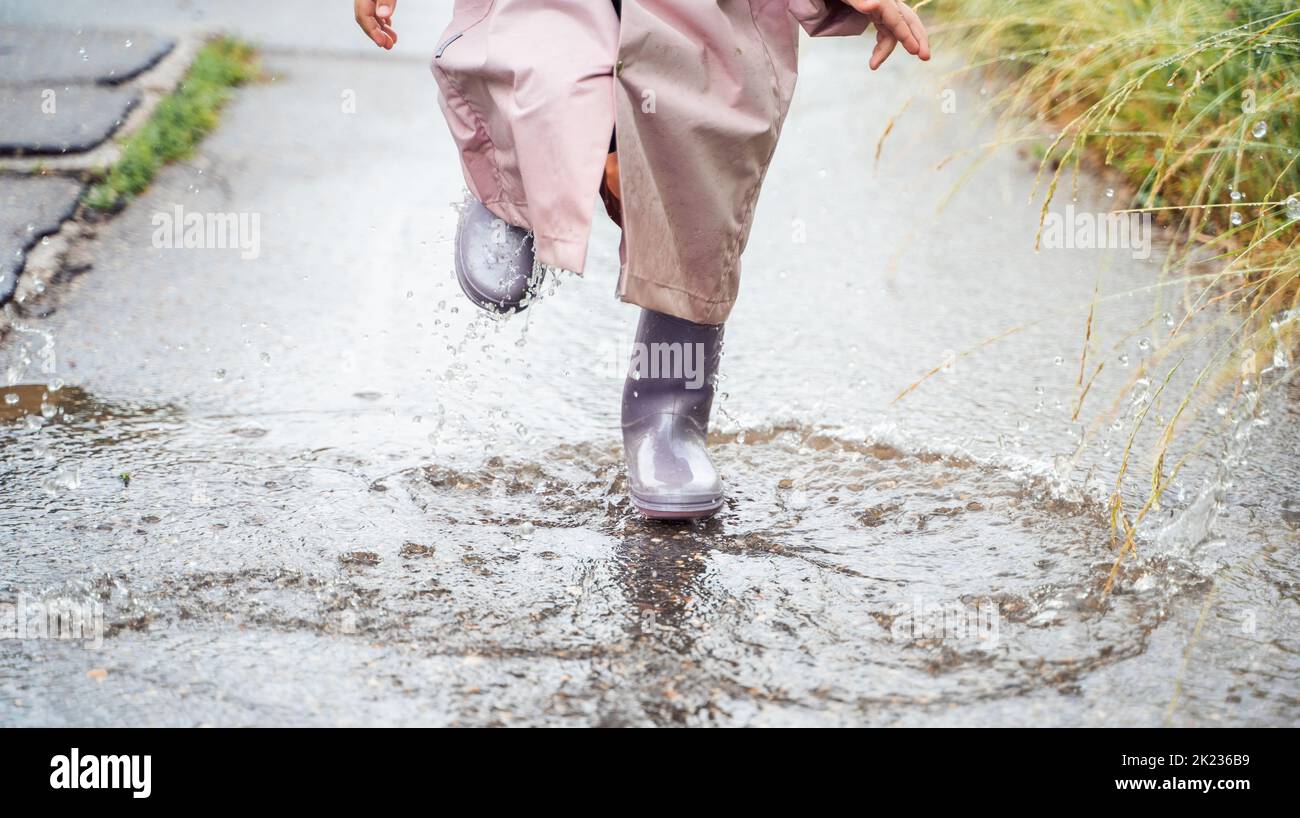 Little girl in pink waterproof raincoat, purple rubber boots funny jumps through puddles on ...