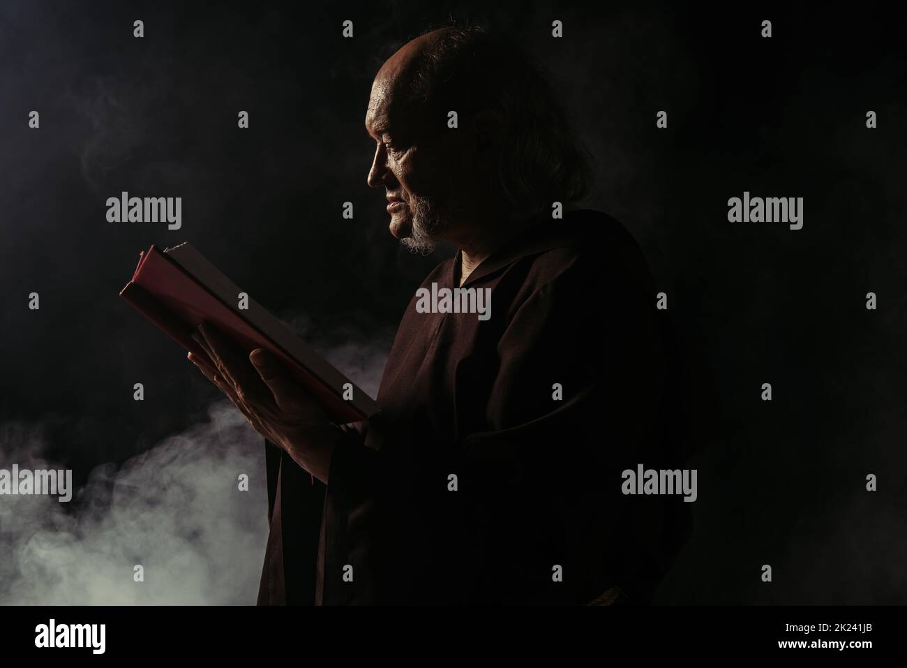 side view of senior priest in cassock reading holy bible on black with ...