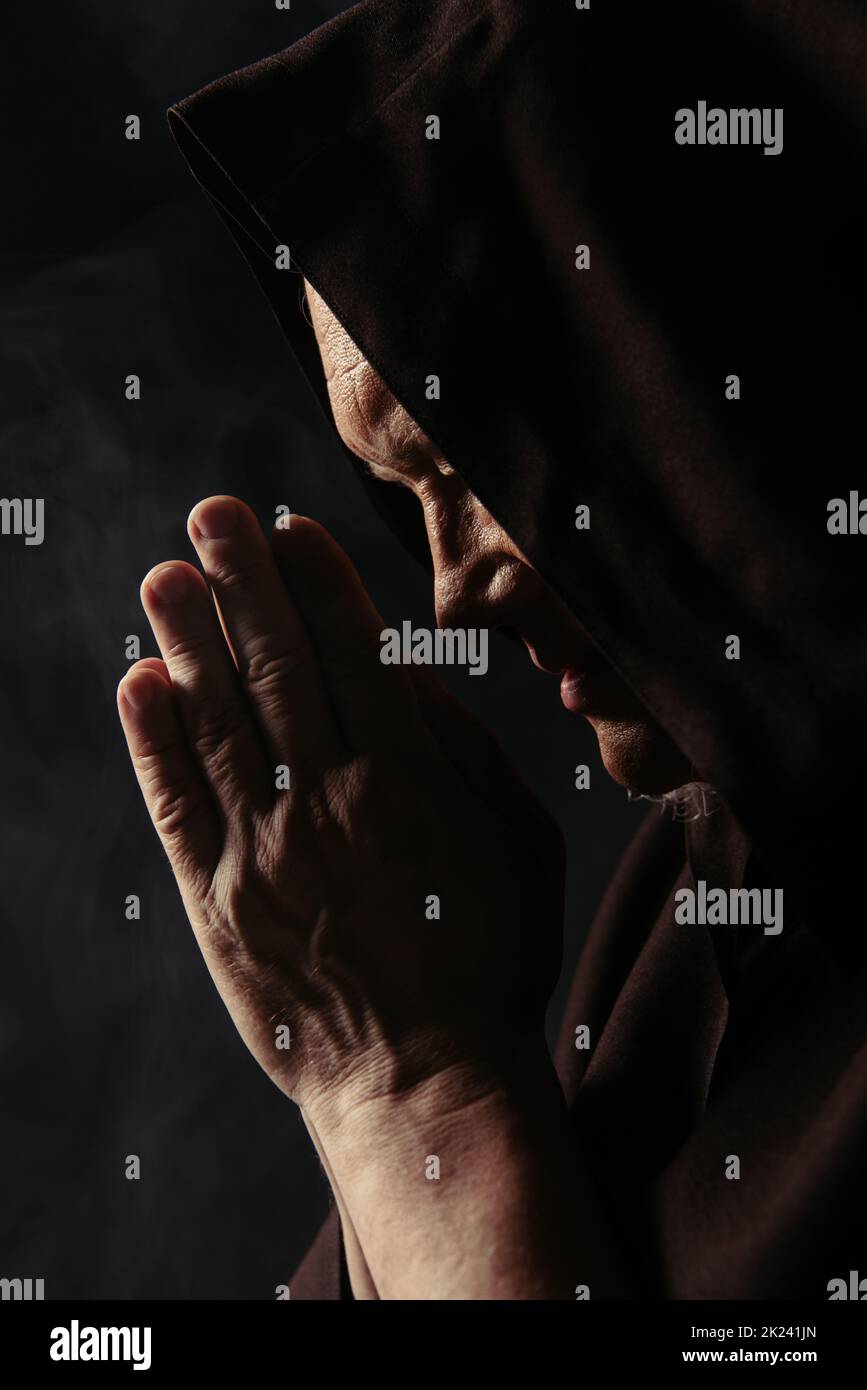side view of senior monk in dark hood and with praying hands isolated ...