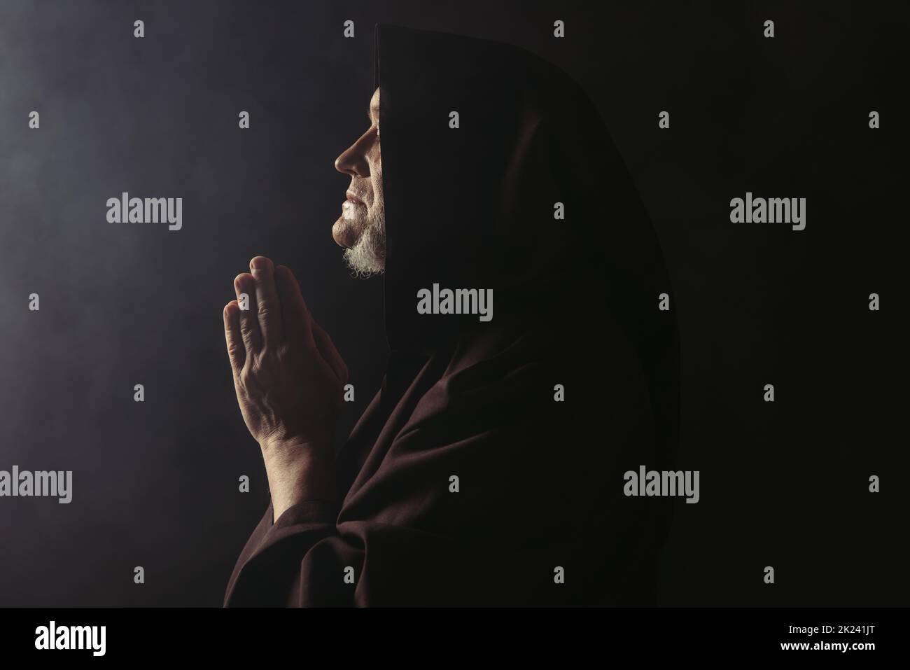 side view of medieval monk in hooded robe praying on black with smoke ...