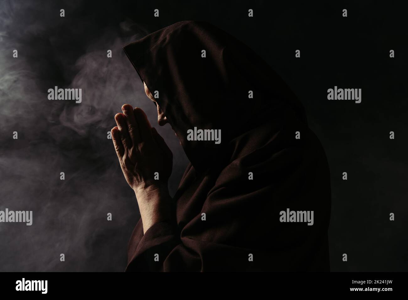 side view of priest with face obscured by hood praying on black with ...