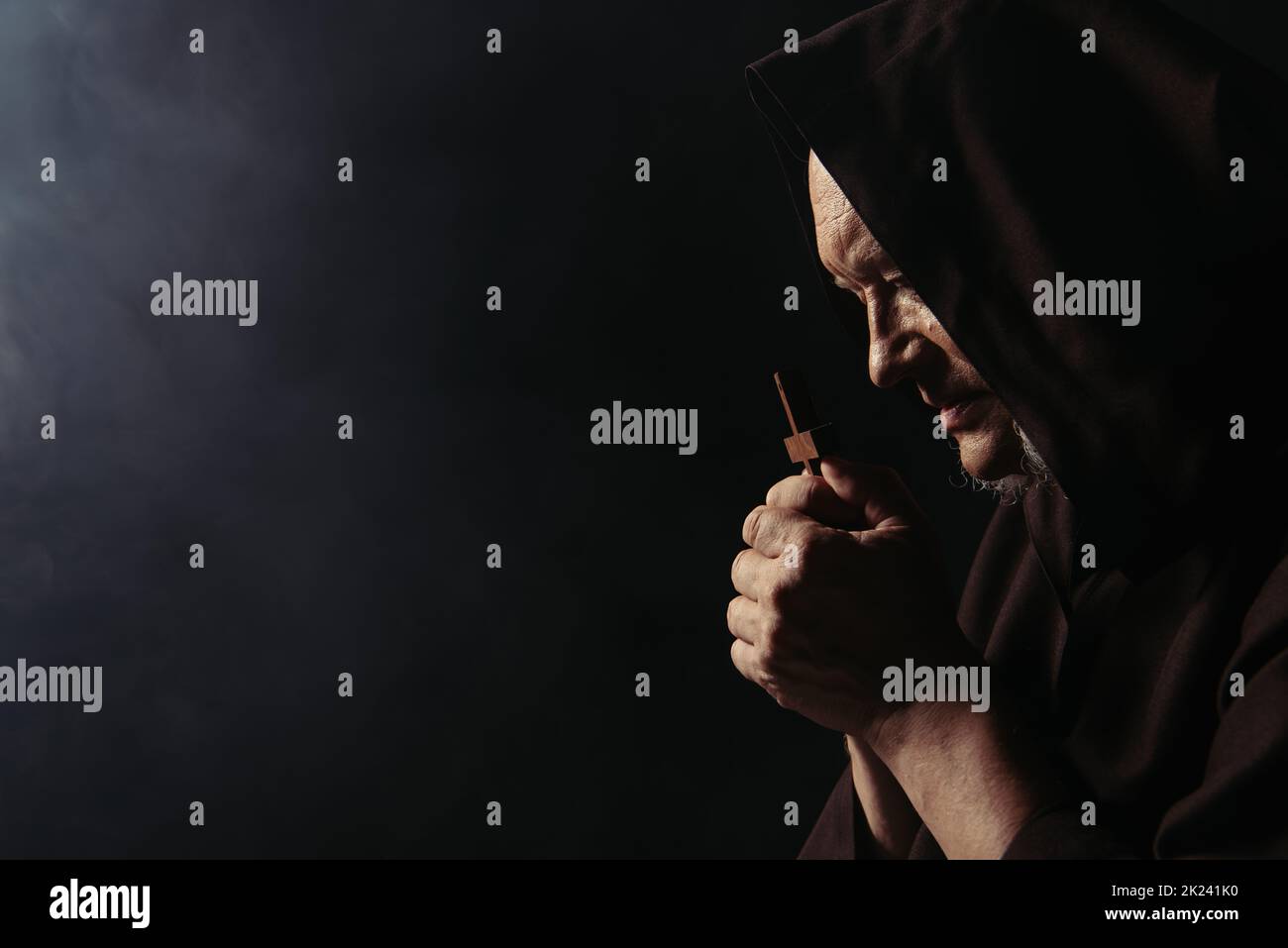 side view of monk in hood praying with holy crucifix on black ...
