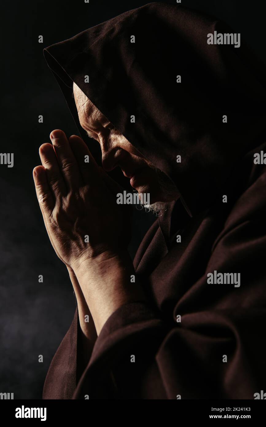 side view of senior priest with praying hands and in black hood on dark ...