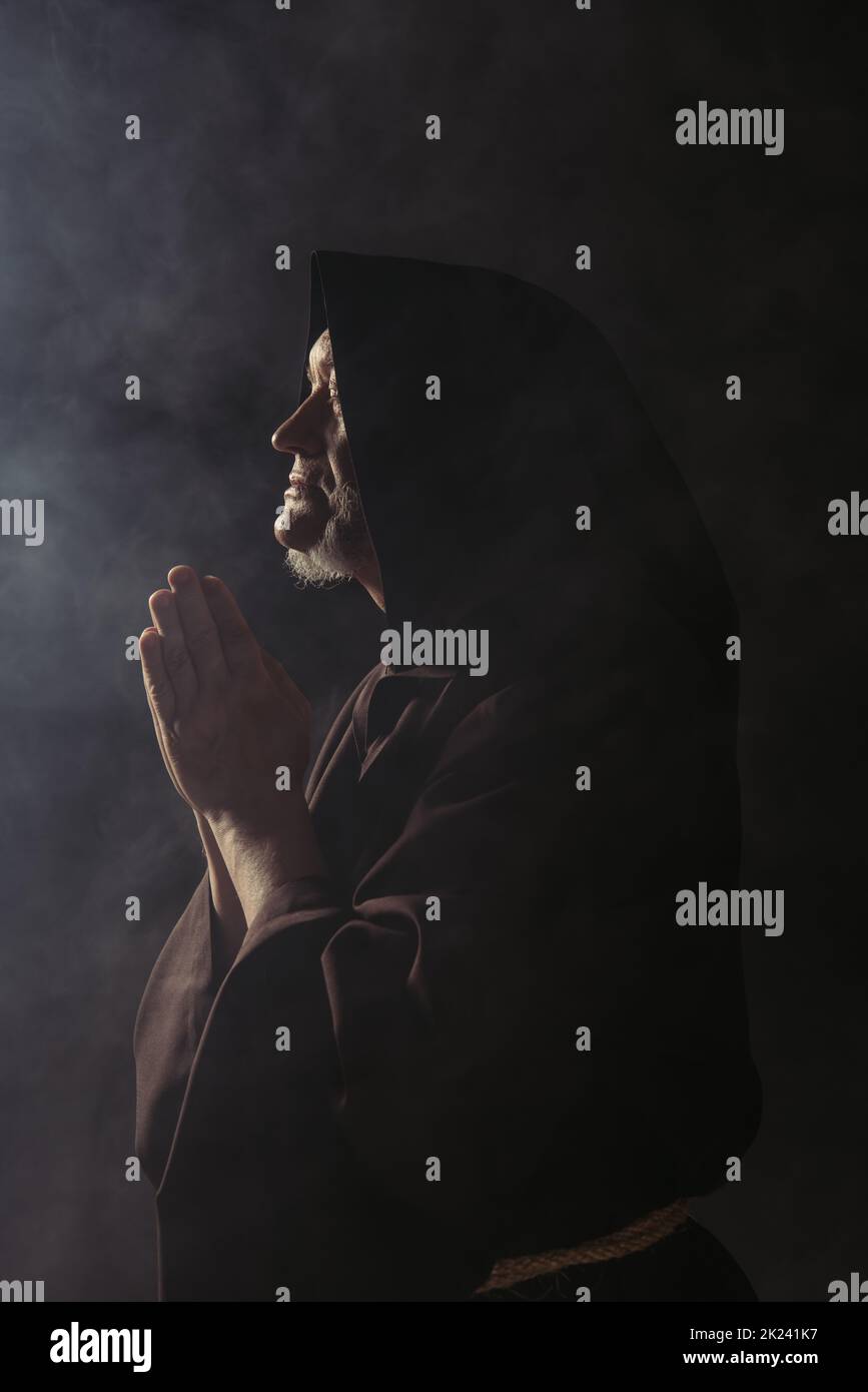 side view of medieval monk in hooded cassock praying on black with ...