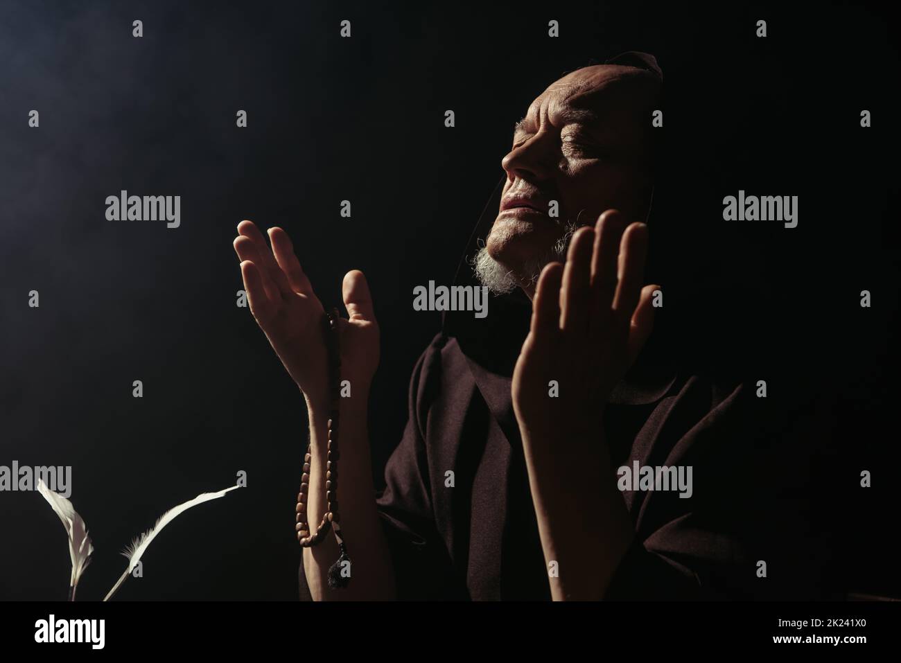 medieval priest praying with rosary and closed eyes isolated on black ...