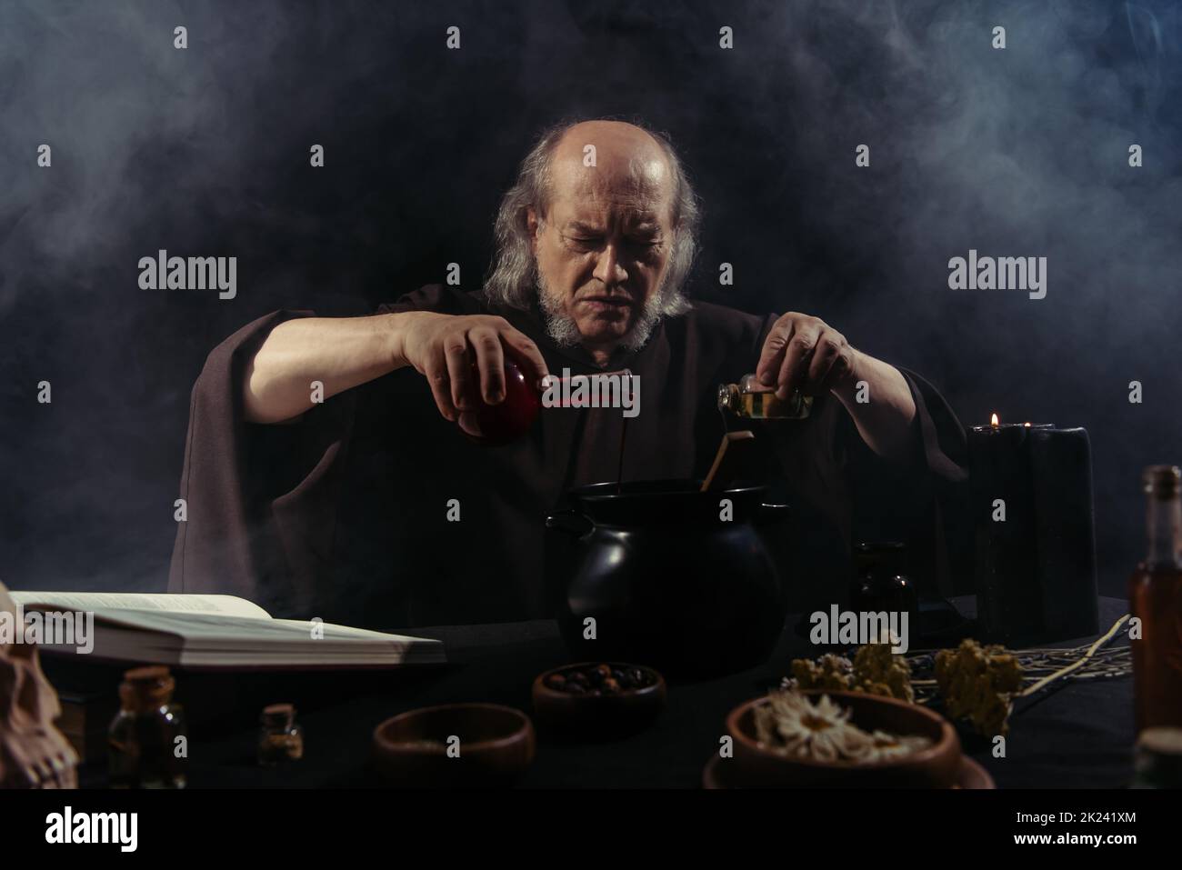 senior magician pouring liquid into pot while preparing potion on black ...