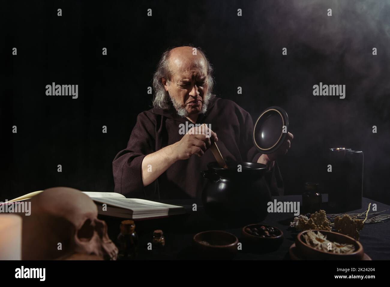 medieval alchemic frowning near steaming pot on black background,stock ...