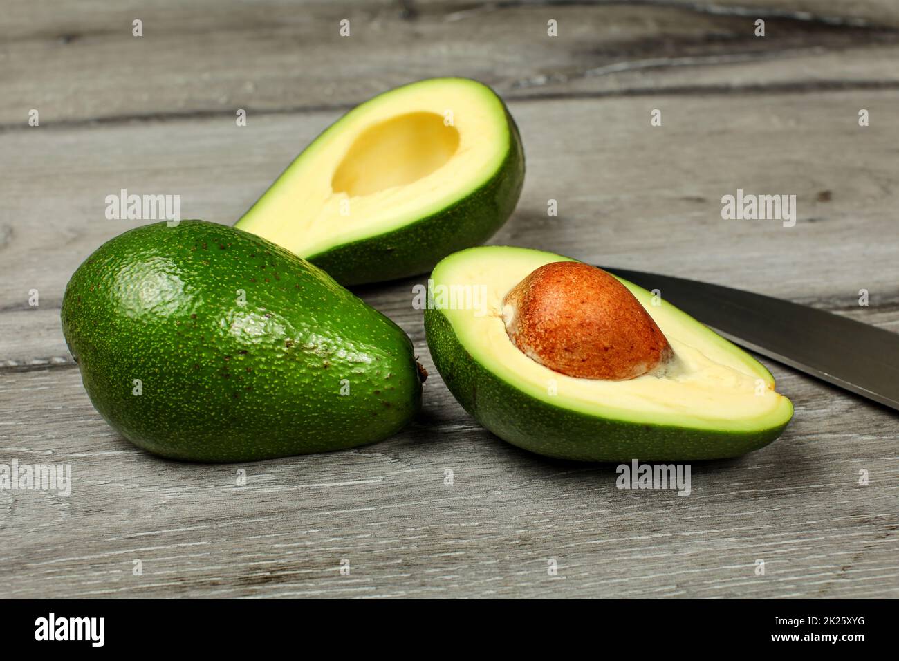 Two avocados, one cut in half, seed visible, knife next to it, on gray ...