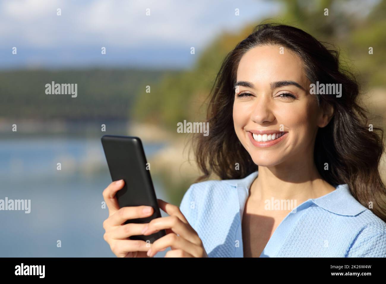 Happy woman holding phone looking at you in nature Stock Photo - Alamy