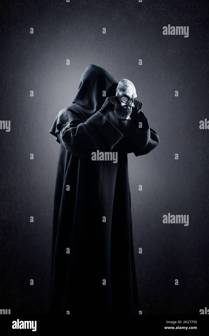 Creepy figure in hooded cloak with human skull mask in hands over dark ...