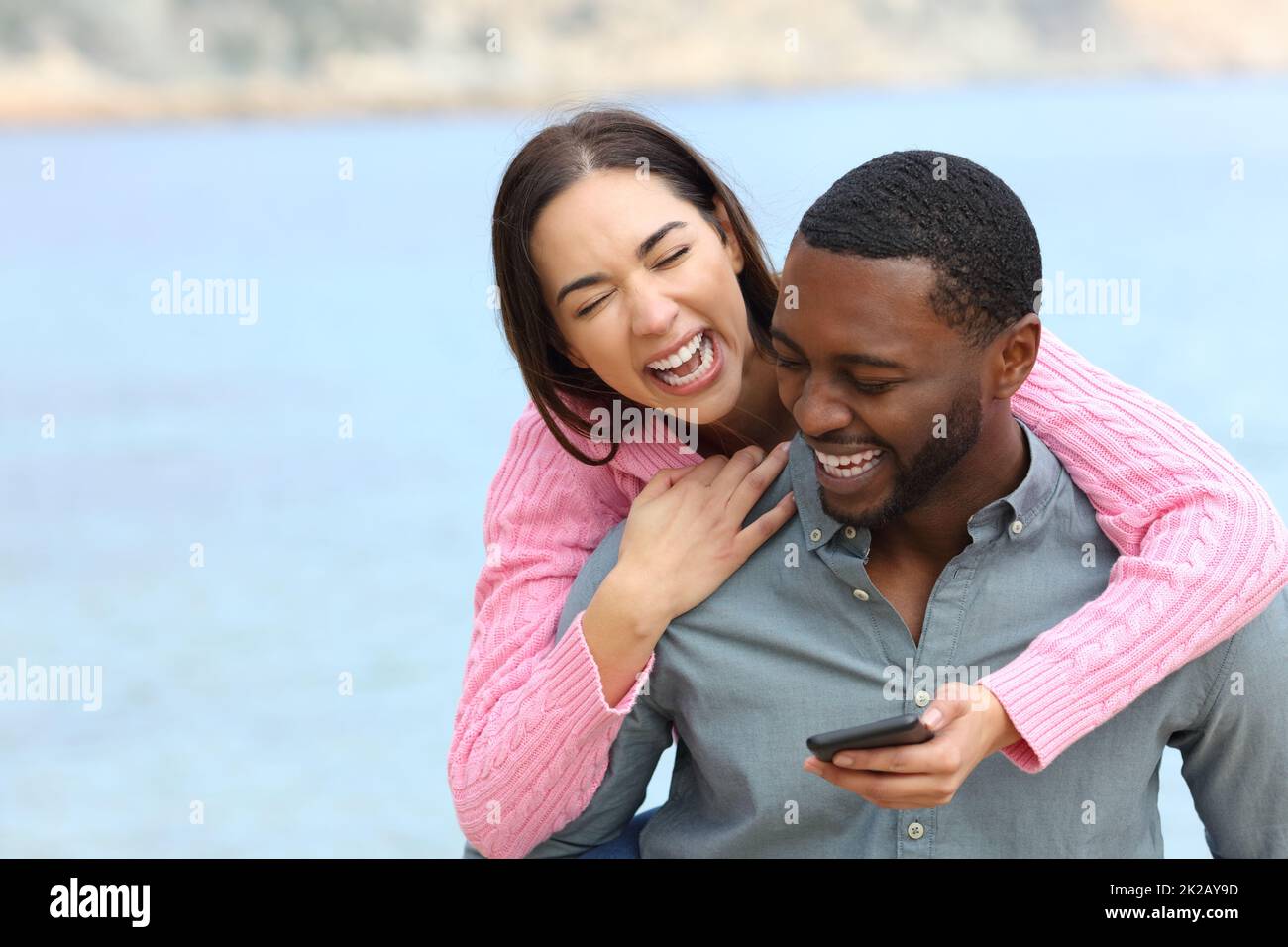 Interracial Couple Laughing Holding Smart Phone On The Beach Stock