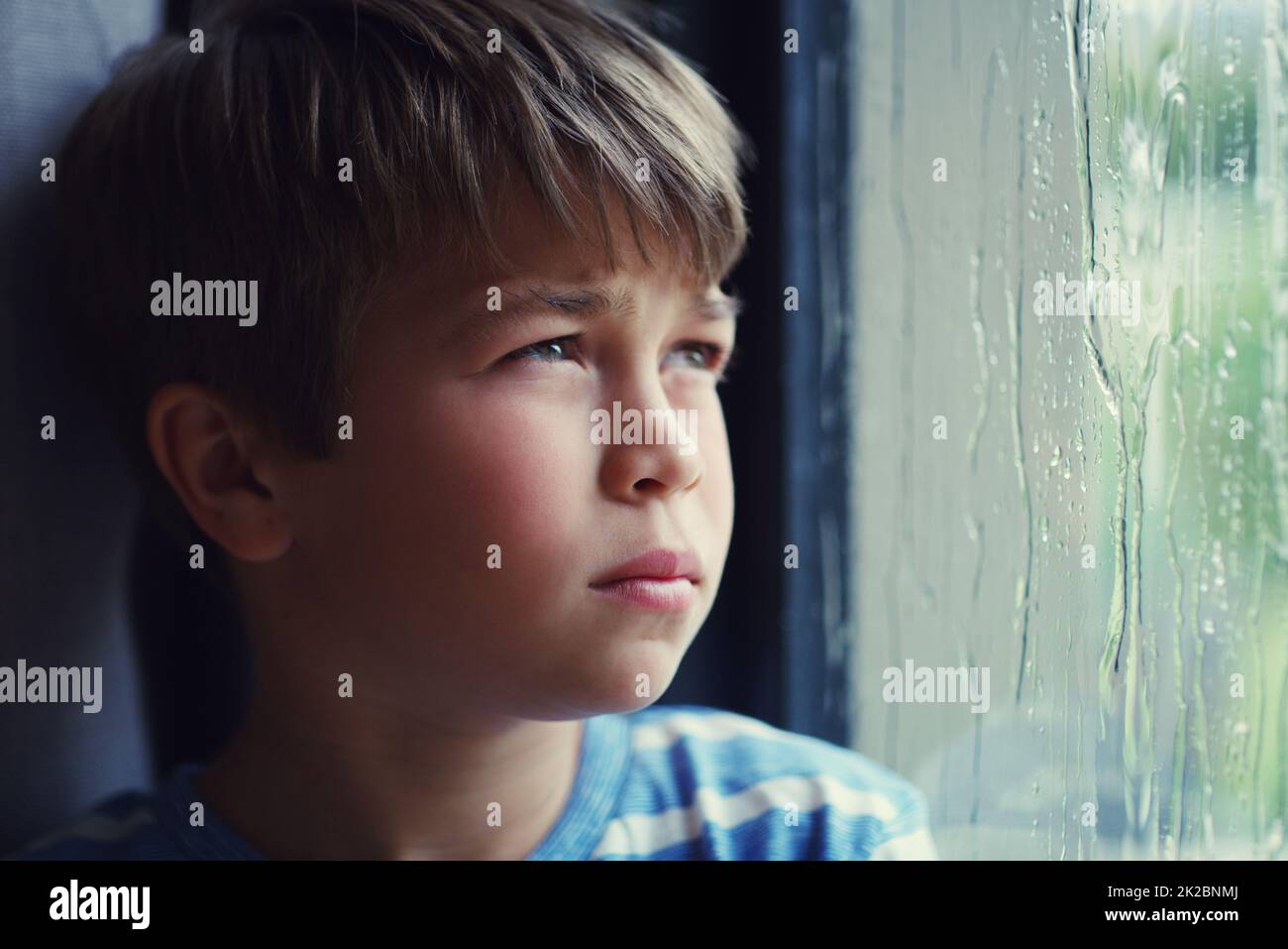 Bored of being bored. Shot of a sad young boy watching the rain through ...