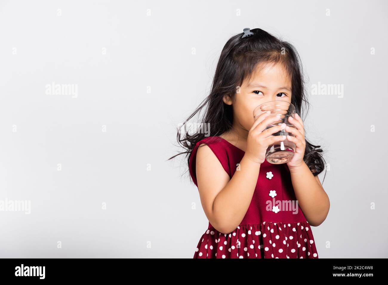 Little cute kid girl 3-4 years old smile drinking fresh water from glass in studio shot isolated ...