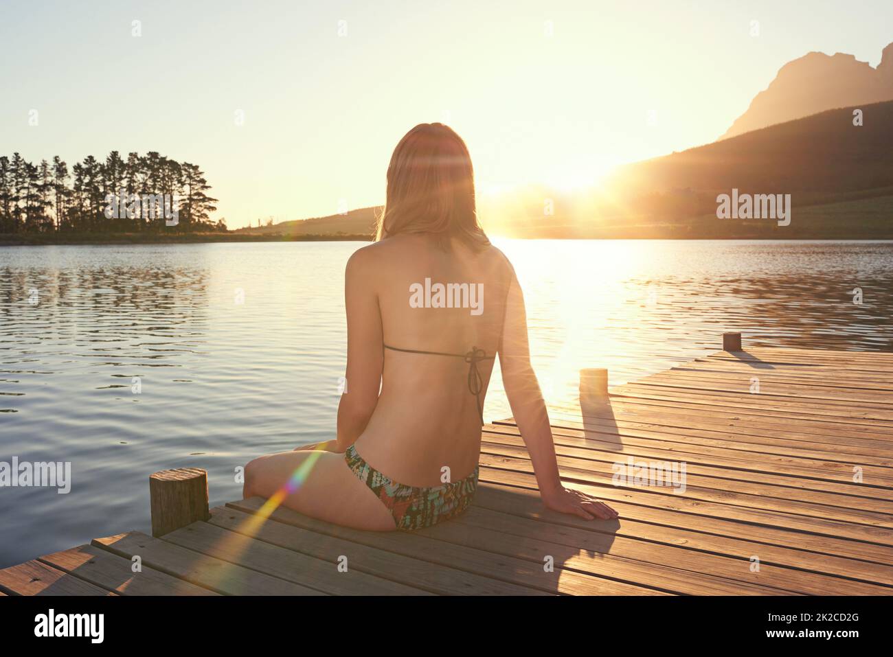 The Lake At Sunset Shot Of A Young Woman In A Bikini Sitting On A Dock By A Lake At Sunset