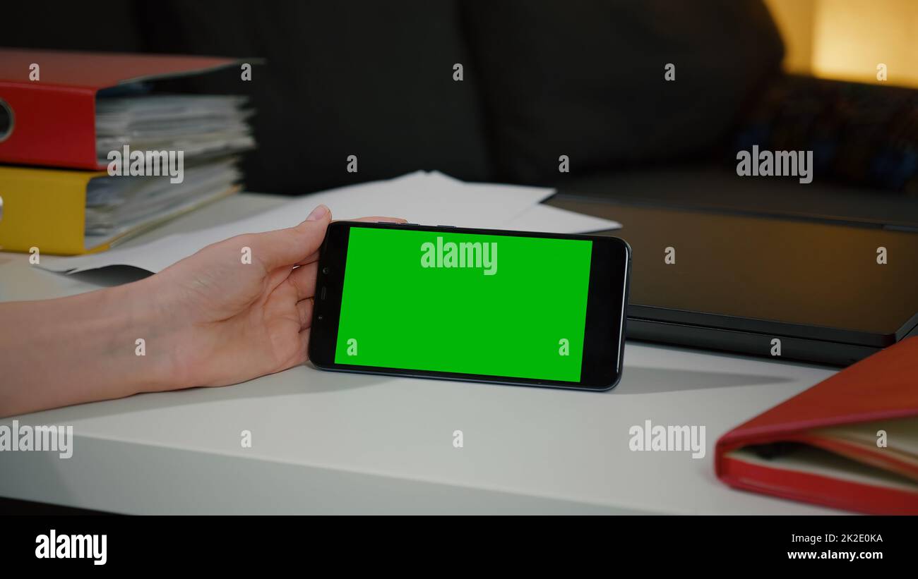 Woman looking at smartphone phone with green screen chroma key display sitting at office desk ...
