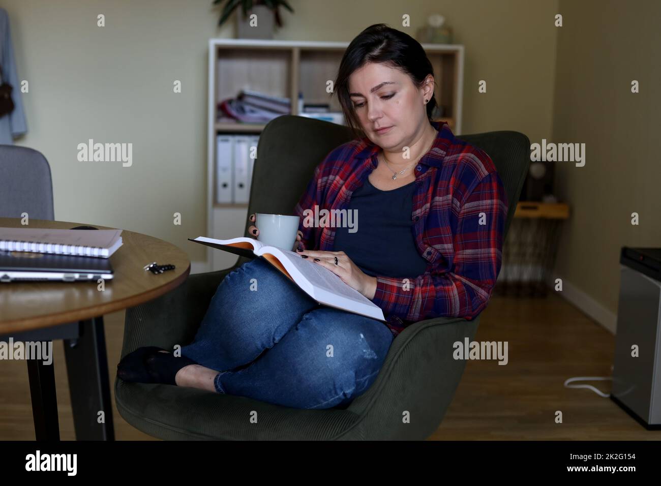 Brunette woman relax with book and cup of tea in armchair at office ...