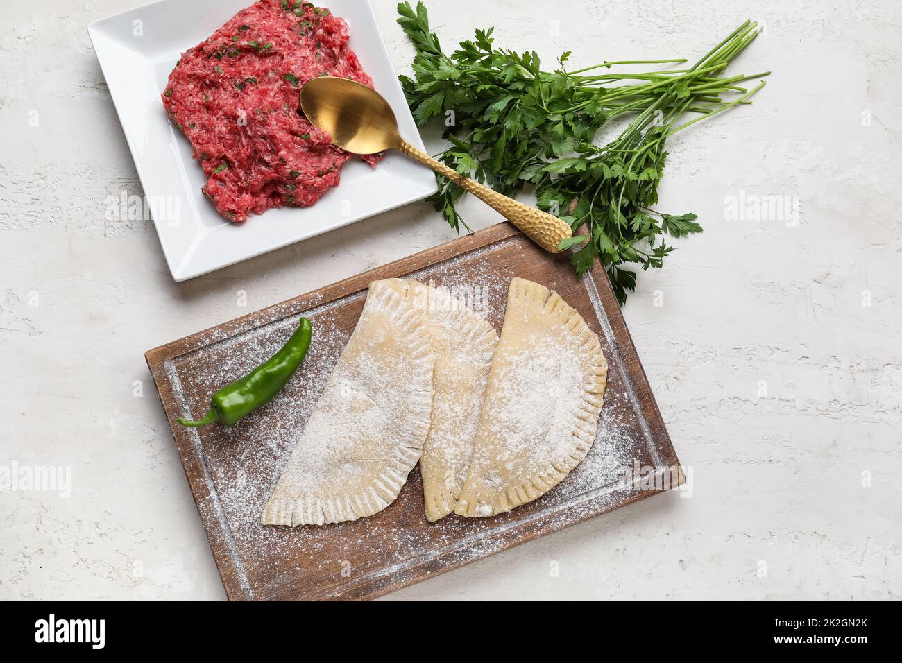 Board with raw chebureks, plate of mince, spoon and parsley on white ...