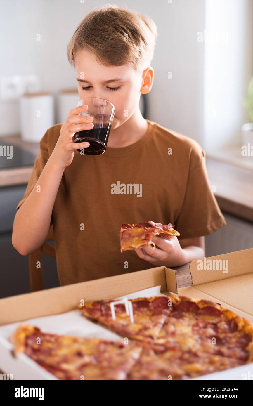 Blond teenager drinks cola and eats pizza in the kitchen at home ...