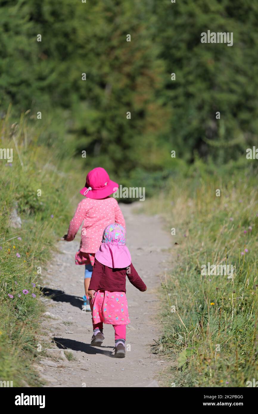 Two toddlers with sun hats are walking up a hiking trail in french alps ...