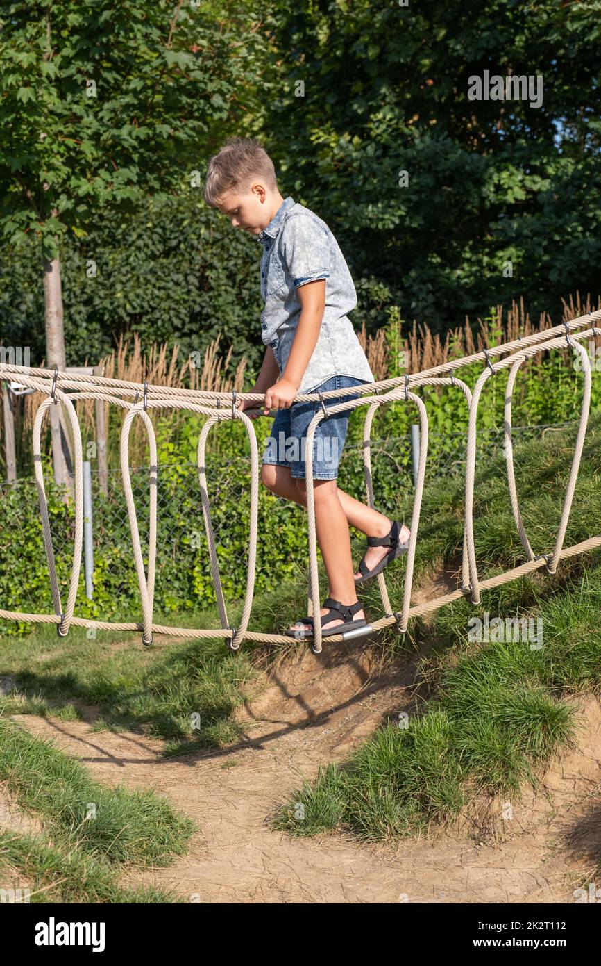 Cheerful blond boy walks on rope bridge on a wooden playground in a ...