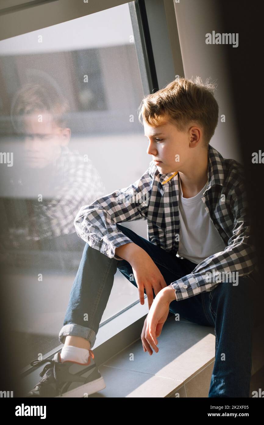 Sad teenager in casual clothes looks out the window while sitting on ...