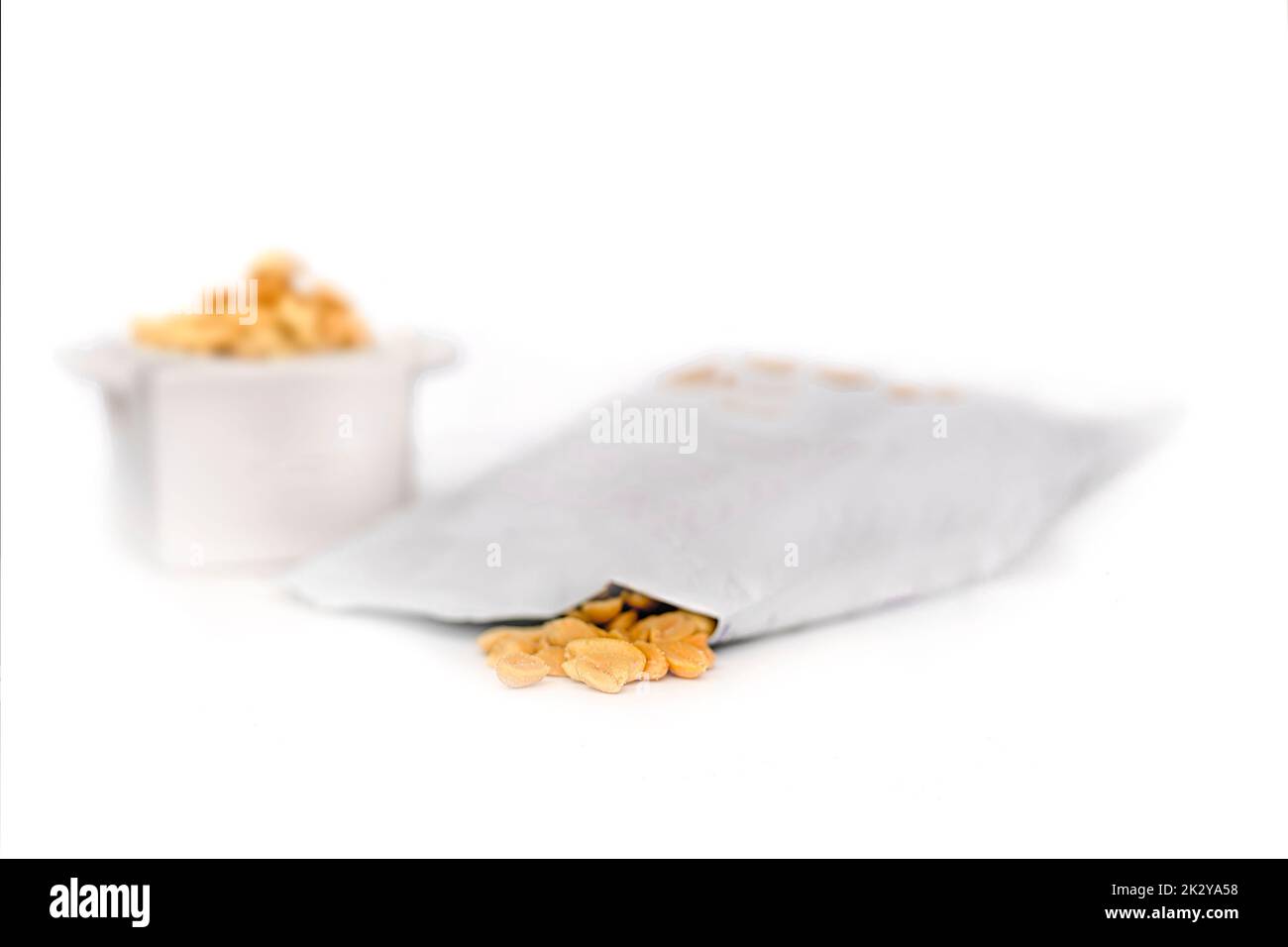 Salted peanuts spilling out of a packet with defocussed pot of peanuts ...