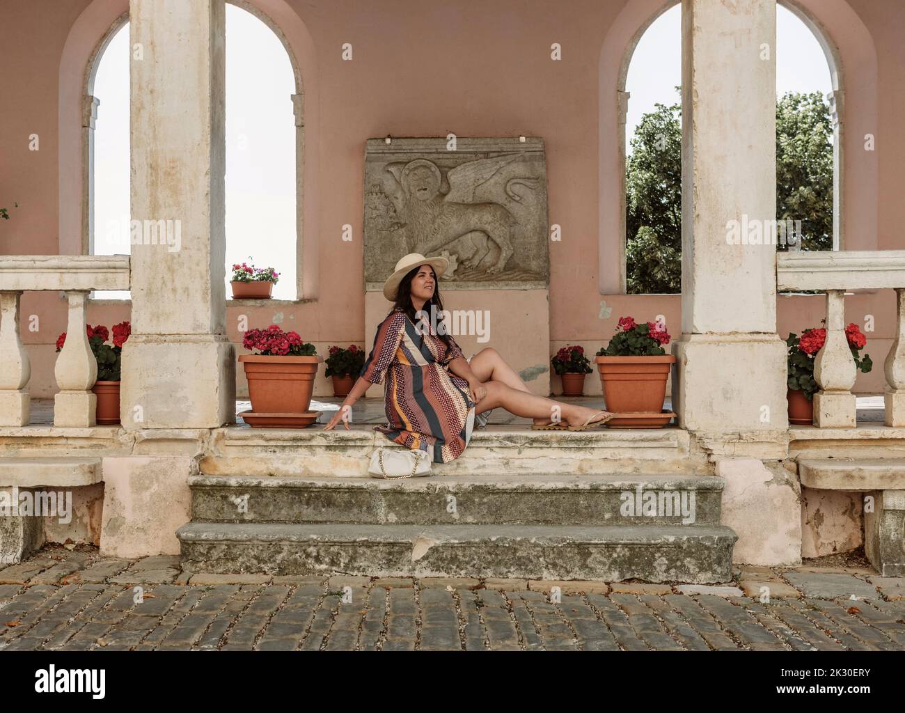 A stylish young woman sitting on stone stairs under arched columns of ...