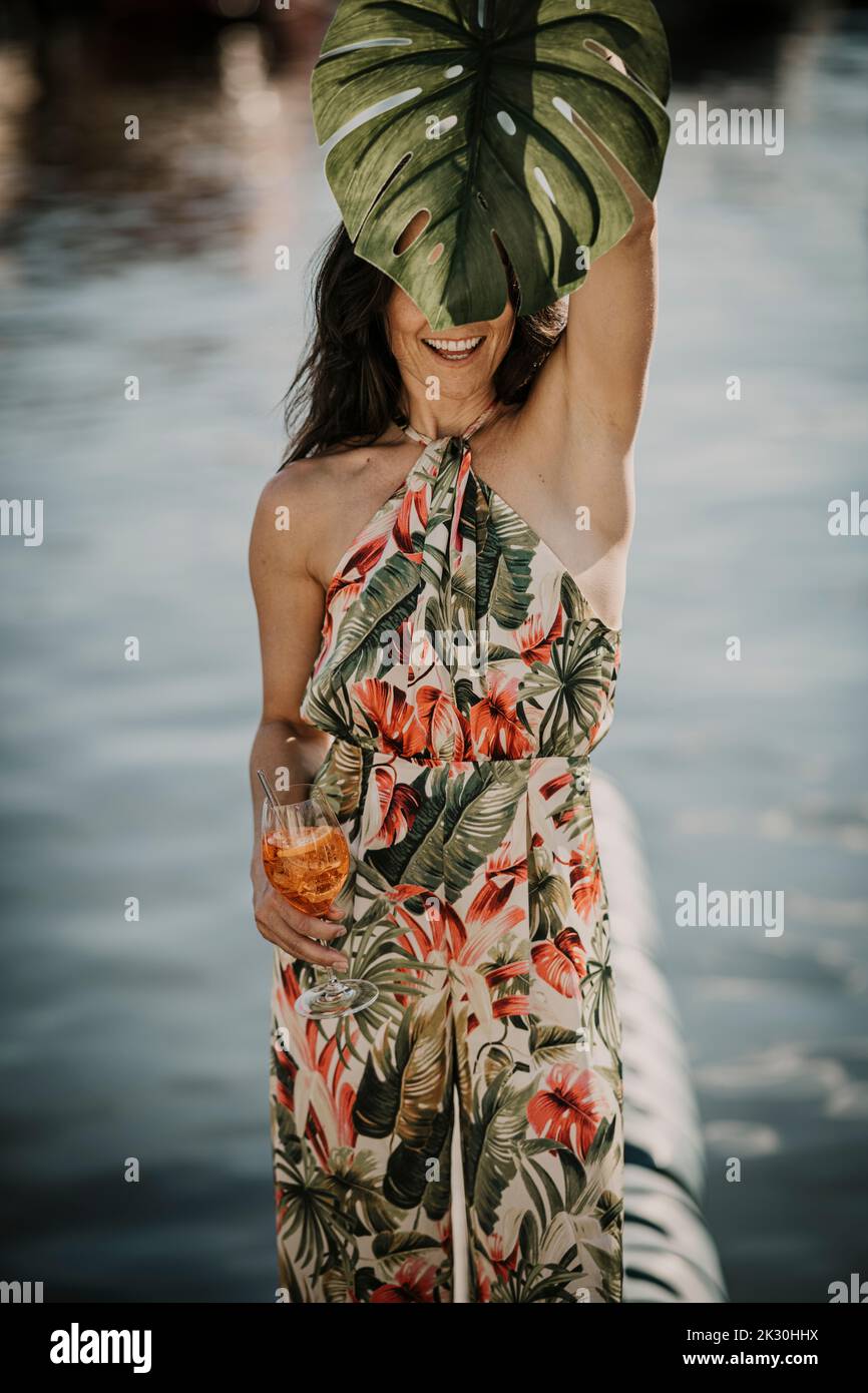 Smiling woman with drink holding monstera leaf Stock Photo - Alamy