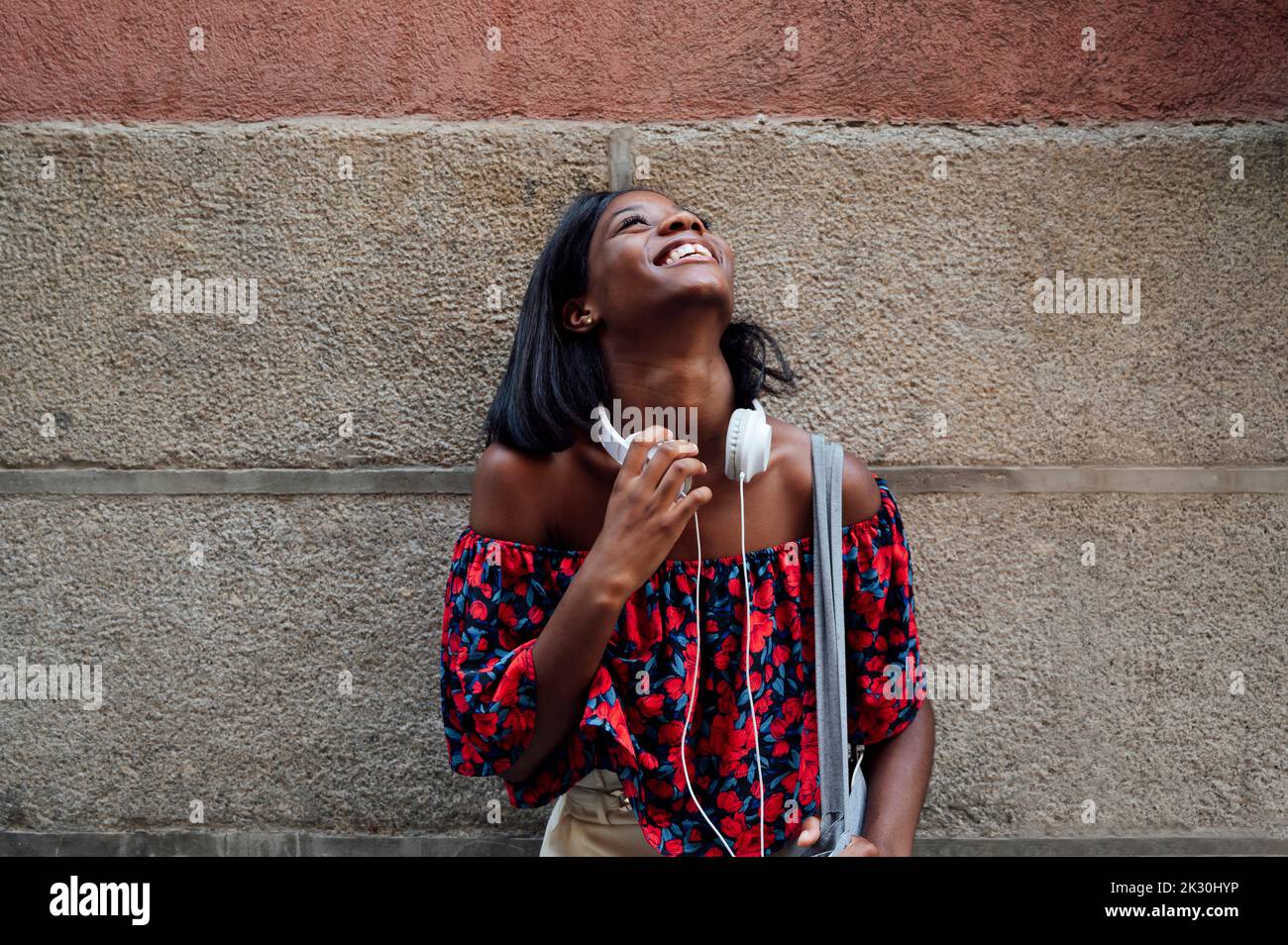 Cheerful woman with headphones looking up in front of wall Stock Photo ...