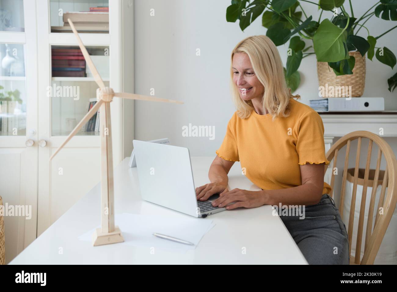 Smiling freelancer with blond hair working on laptop at desk in home ...