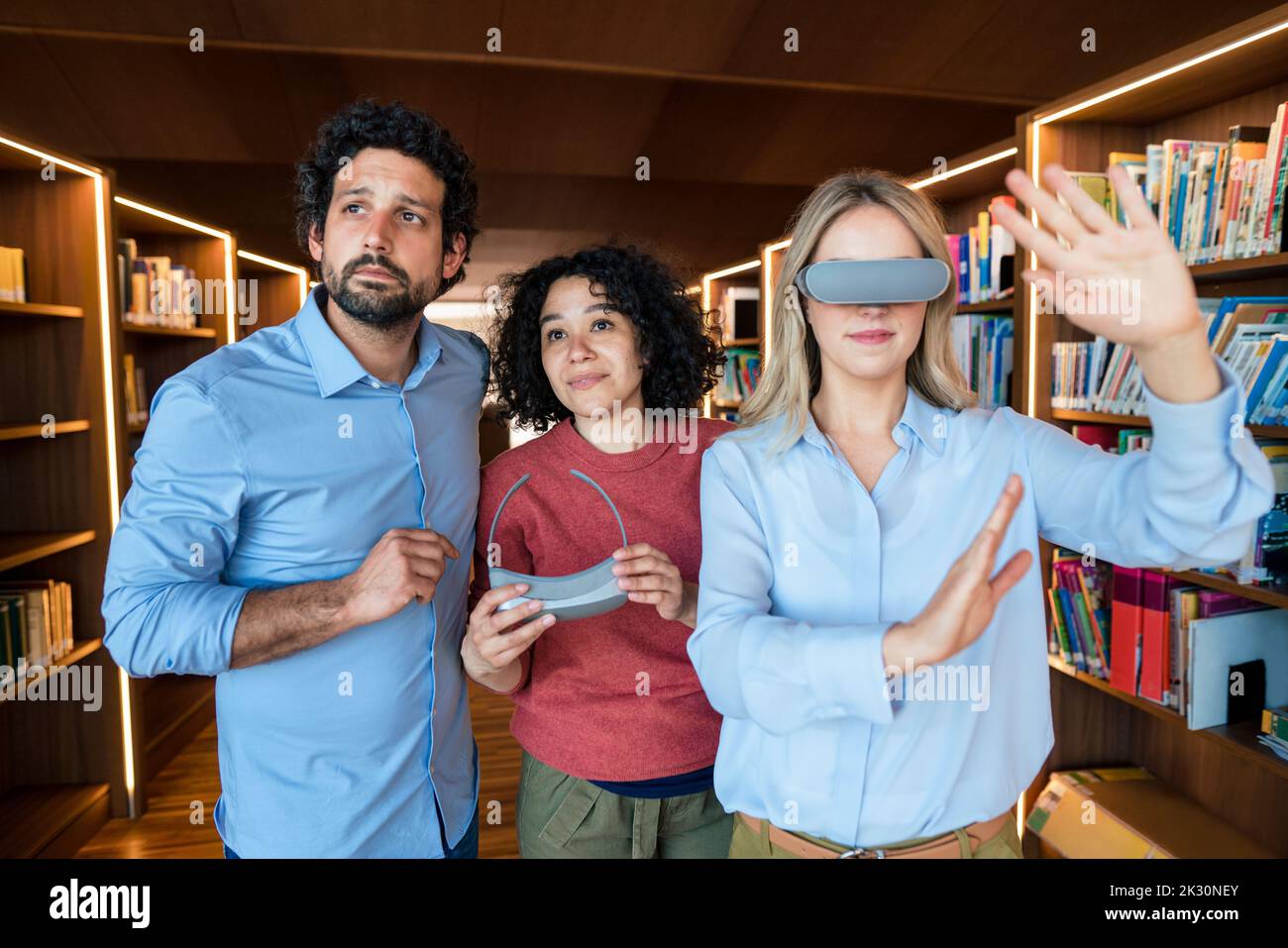 Woman with VR glasses gesturing by colleagues in illuminated library ...