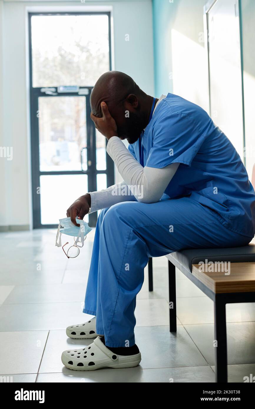 Tired doctor with head in hand sitting on bench in hospital corridor ...