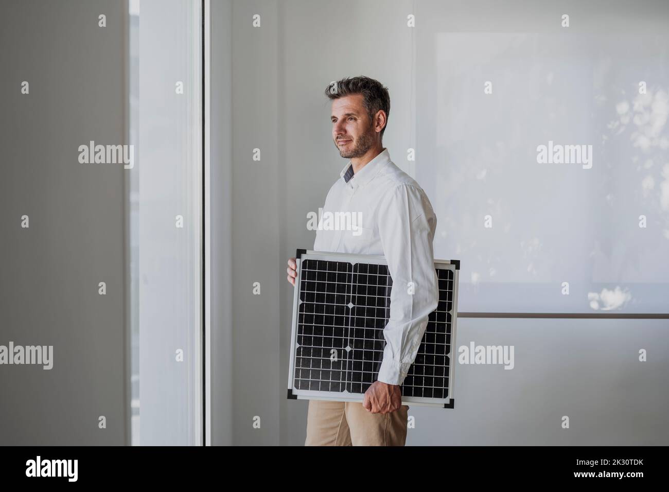Smiling engineer holding solar panel looking through window in office ...