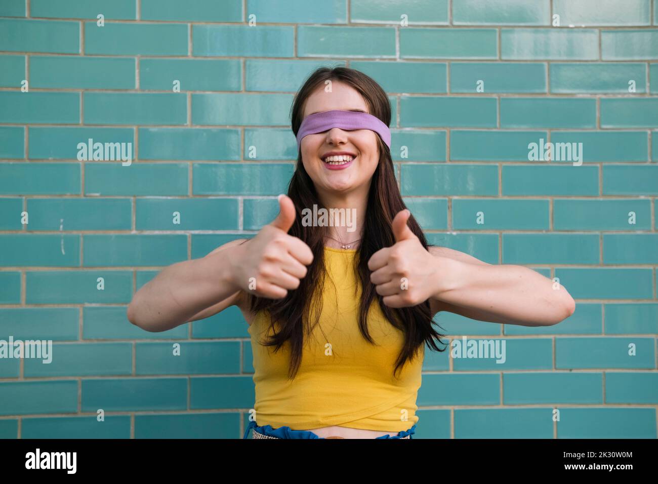 Smiling woman with blindfold showing thumbs up gesture in front of teal ...