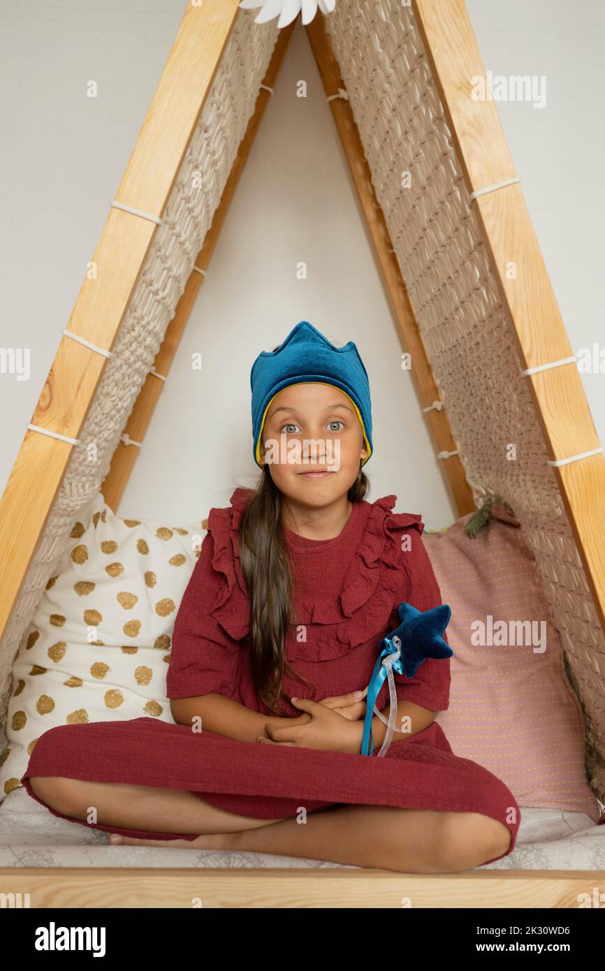Cute girl wearing crown holding magic wand sitting in tent at home ...