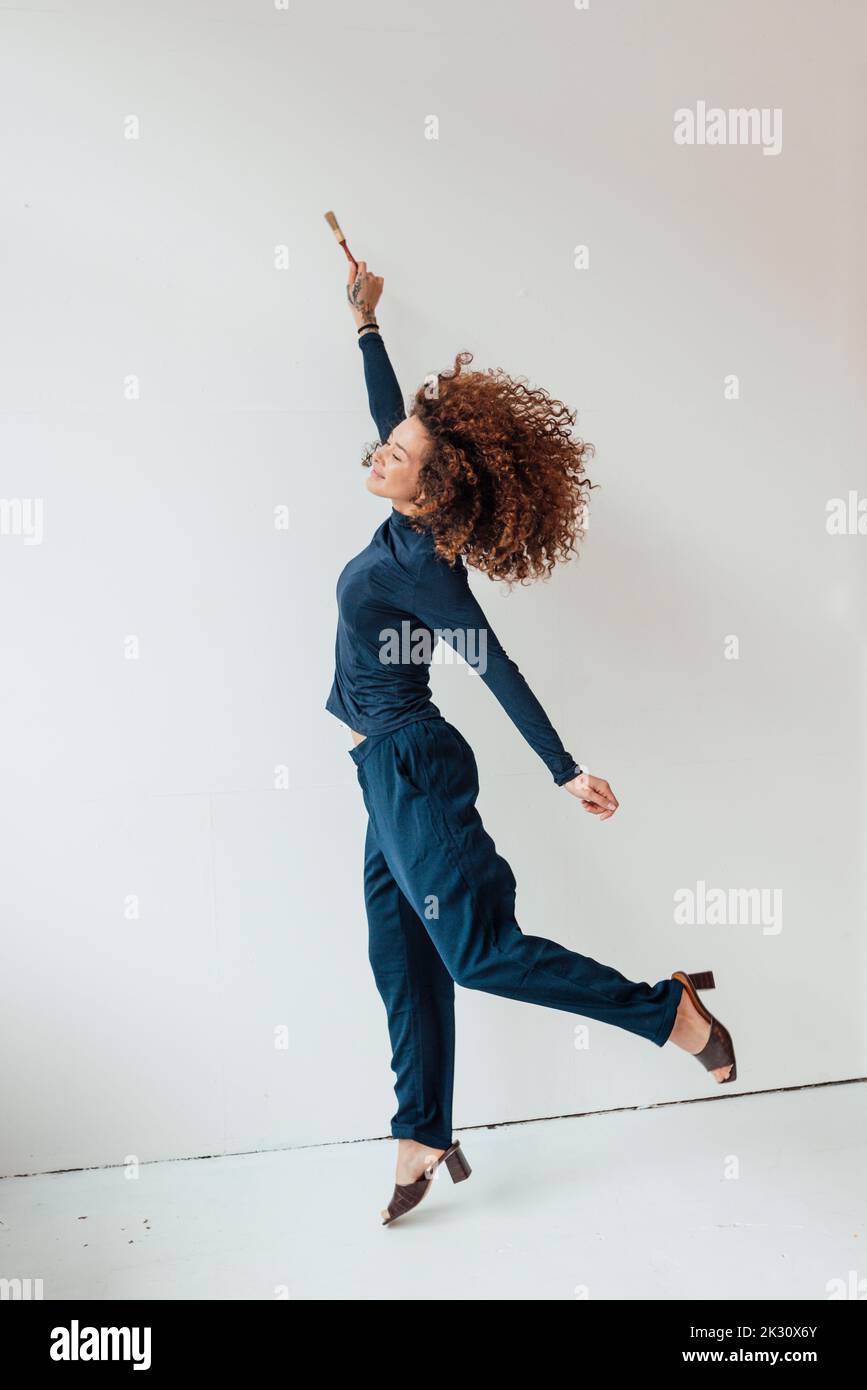Cheerful young woman with hand raised jumping in front of white wall ...