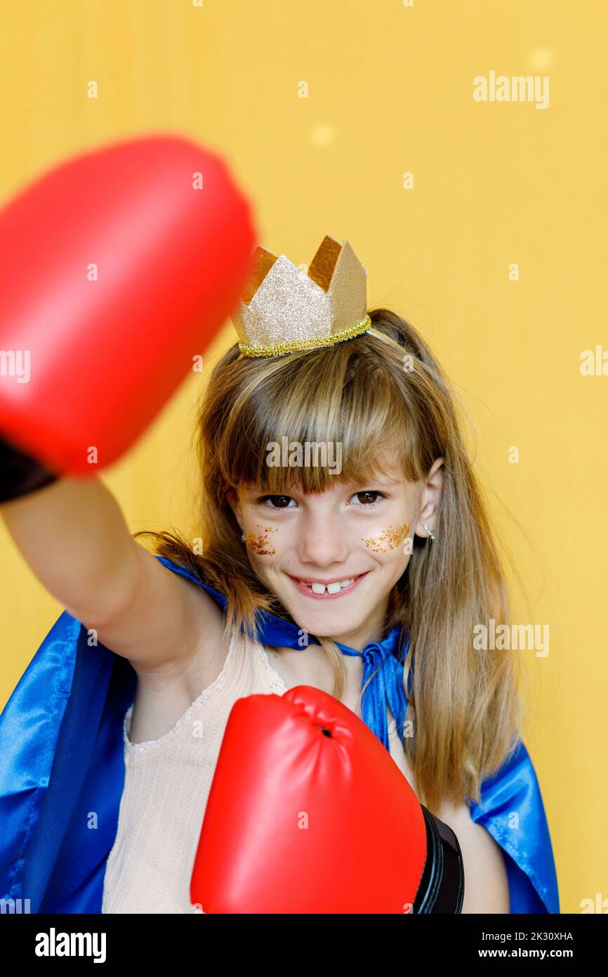 Cheerful girl wearing boxing gloves and crown against yellow background ...
